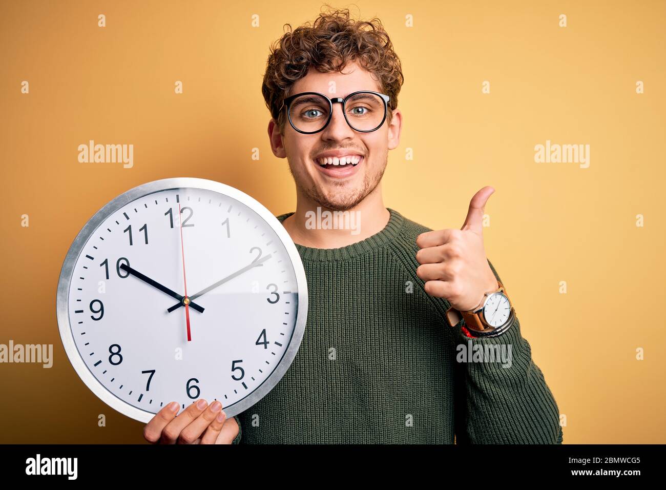 Young blond man with curly hair wearing glasses holding big clock over ...