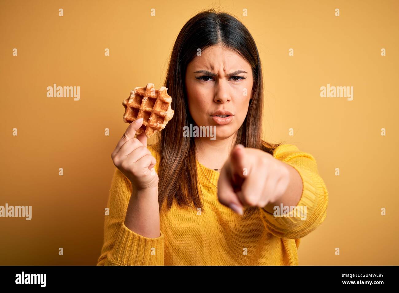 Young beautiful woman eating sweet waffle pastry over yellow background ...