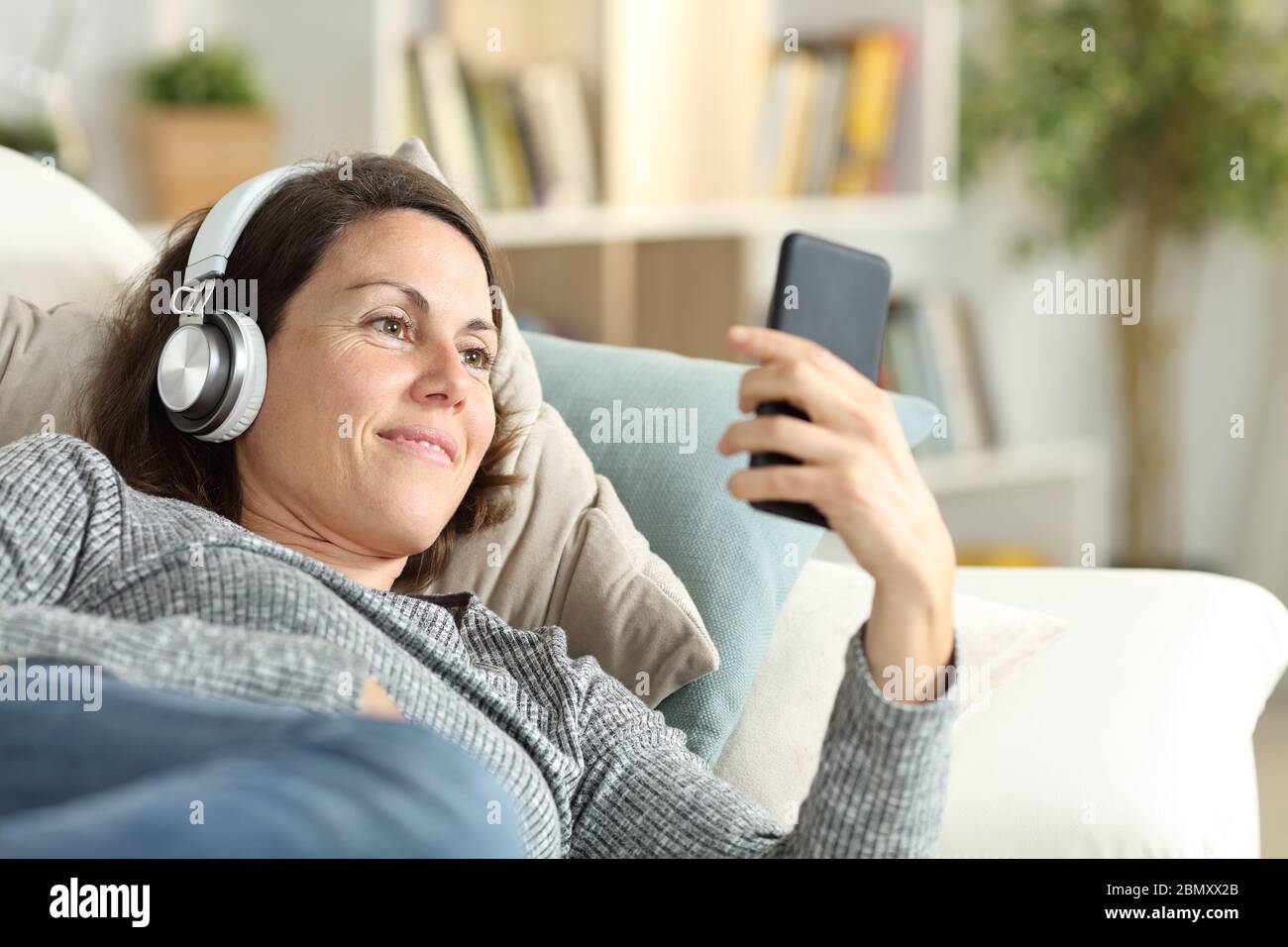 Happy adult lady with headphones listens music on smart phone lying on ...