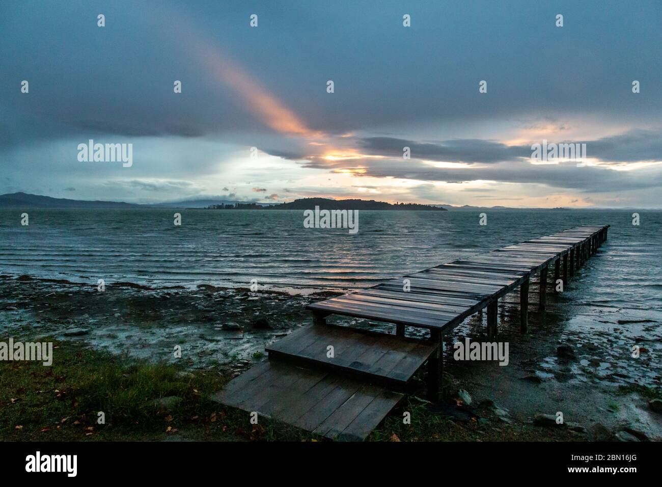 Low point view of a pier on a lake with a ray of sun against a moody ...