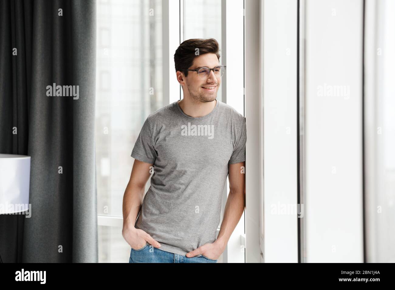 Image of a positive smiling young man indoors at home looking at window ...