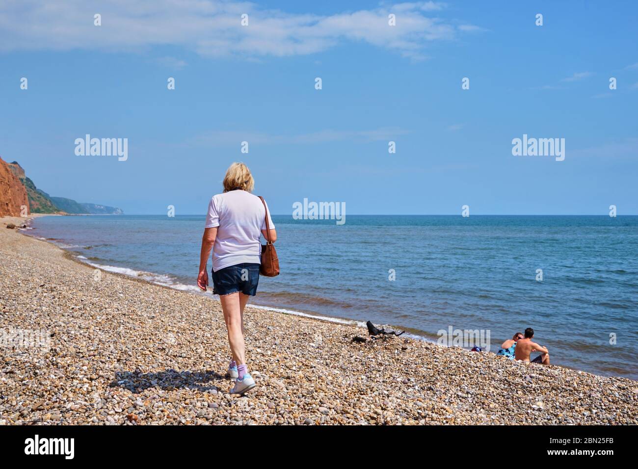 Fair haired woman walking on a pebble beach with 2 other people sitting ...