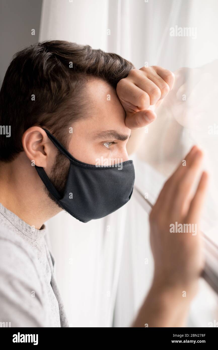 Sad young male patient in facial mask looking out window and leaning on ...