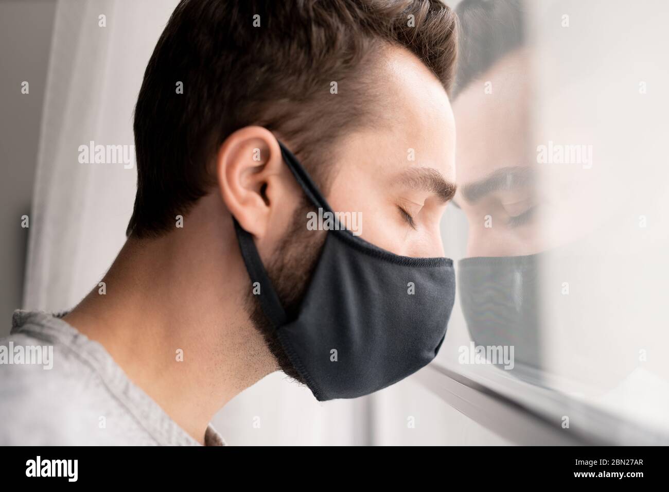 Sad puzzled young man in cloth mask leaning head on window and keeping ...