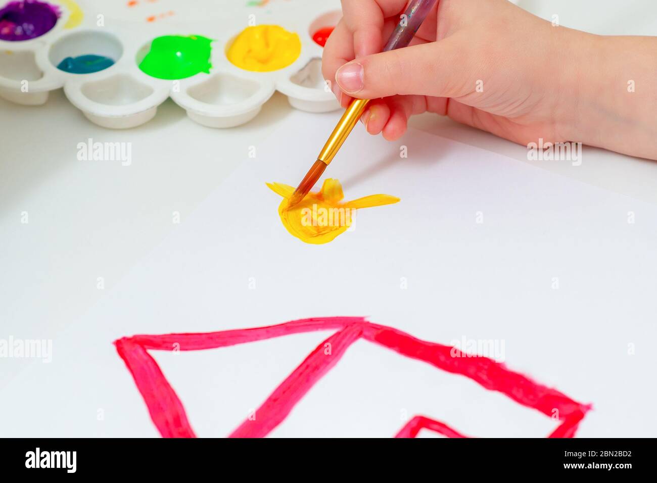 Hand of child drawing yellow sun with red house with brush and ...