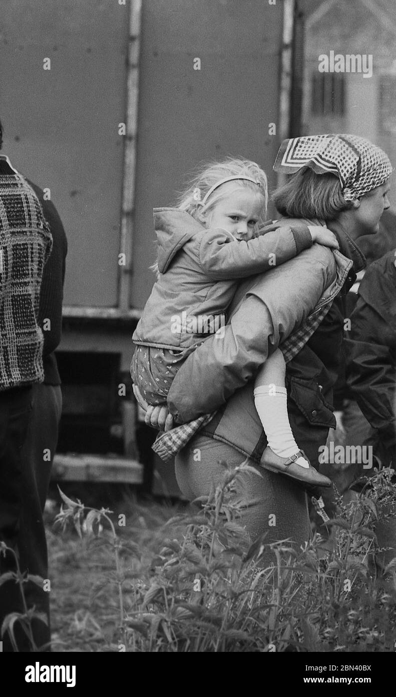 1980s, outside at a country fair, a mother carrying her tired young ...