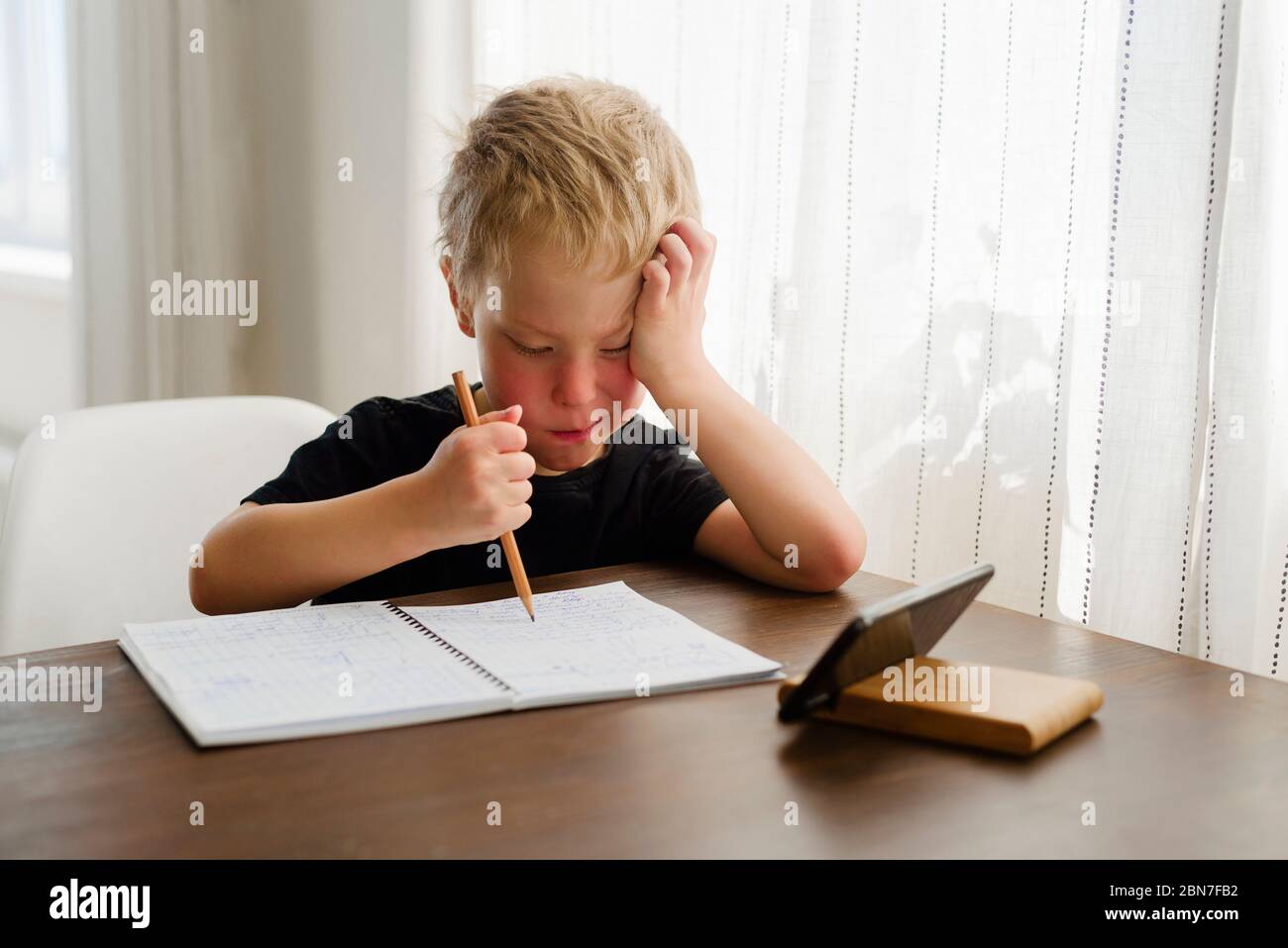 Little sad and tired boy learning at home. Watching webinar by mobile ...
