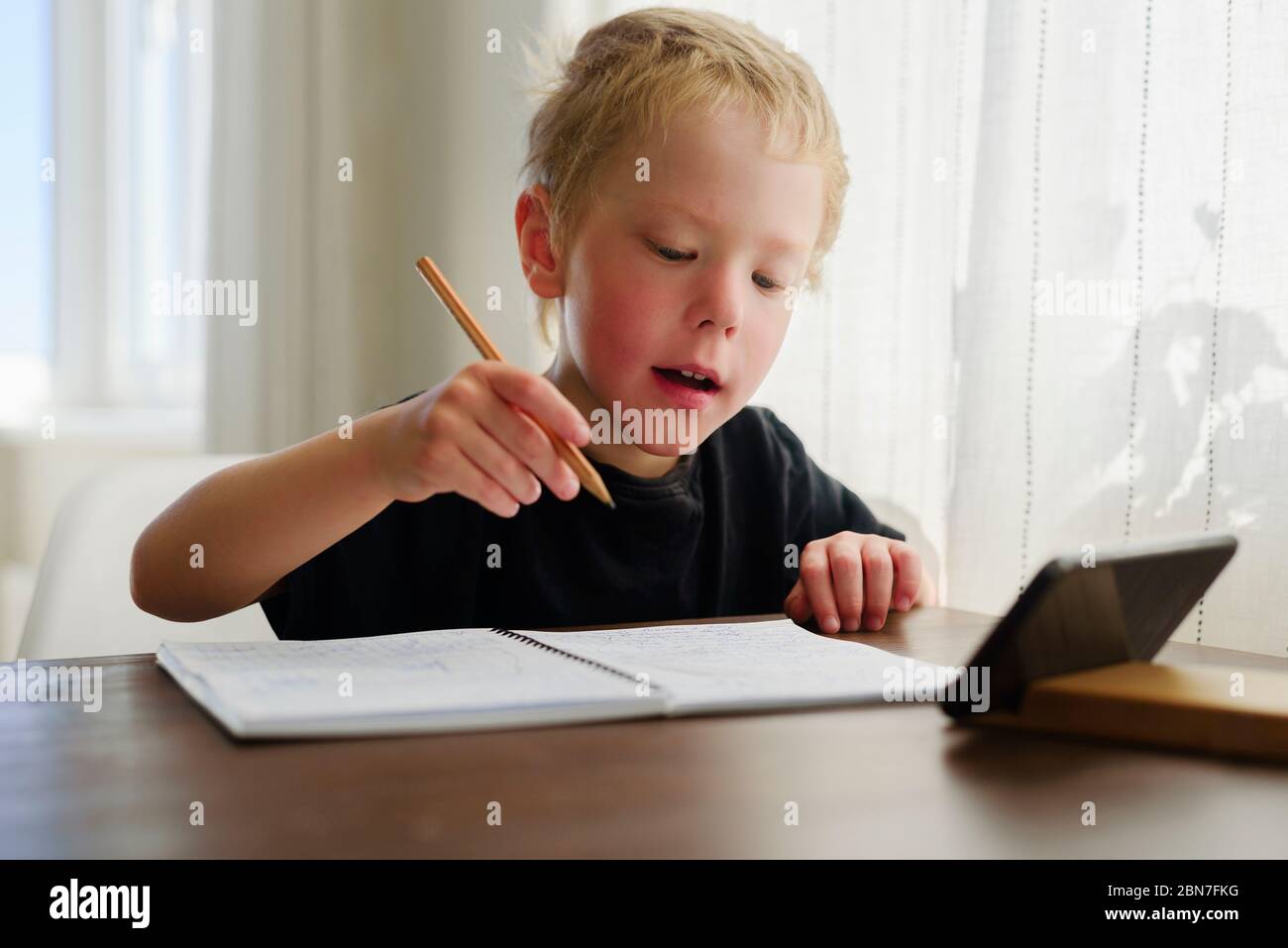 Little boy learning at home. Watching webinar by mobile, writing in ...