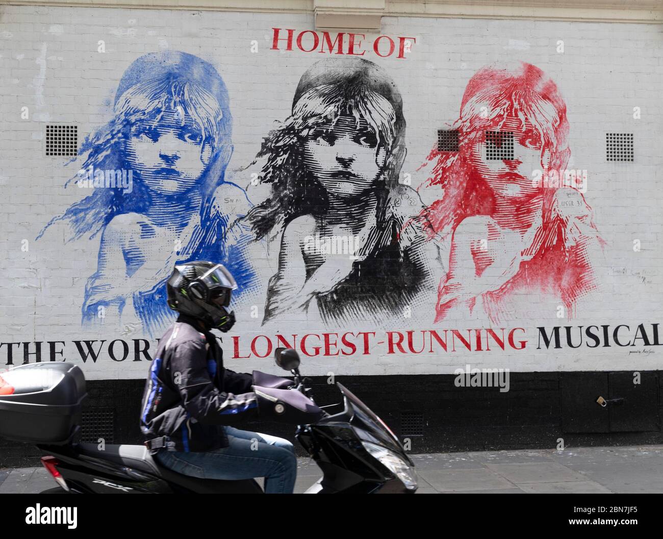 London, UK. 12th May, 2020. A man rides a motorbike past a poster of ...