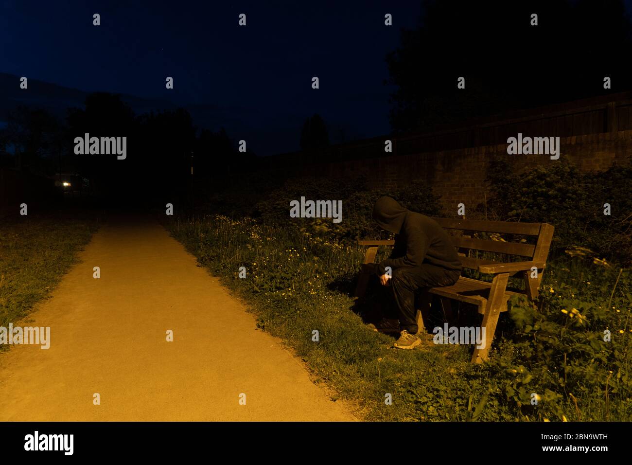 A mysterious hooded figure sitting on a bench, lit up by a street light ...