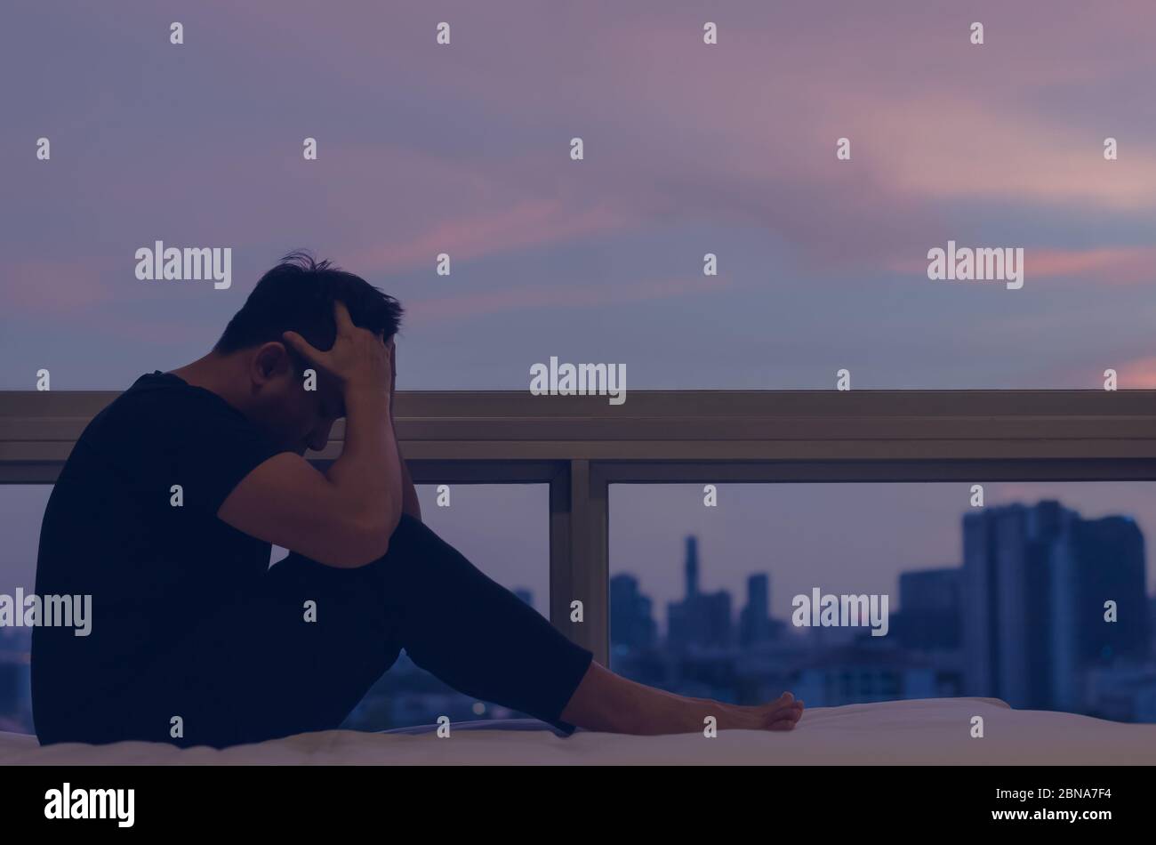 A person sitting and feeling depressed on bed with city view in dusk ...