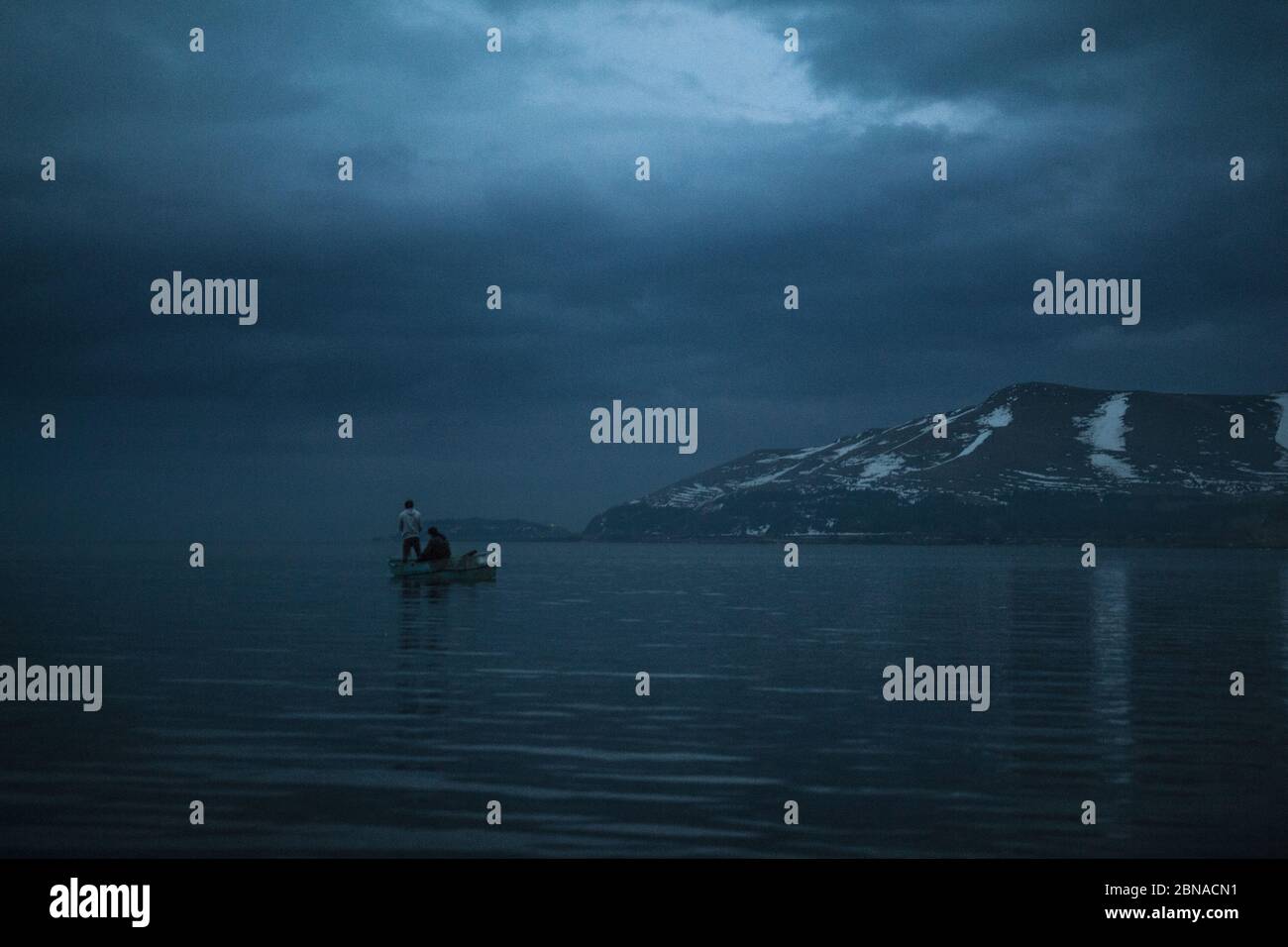 Eerie shot of two males in a small boat on the sea with mountain ...