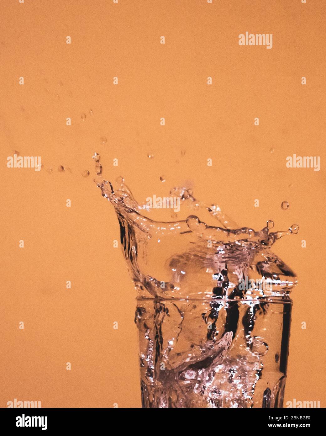 Vertical shot of water splashing out of a glass on an orange background ...