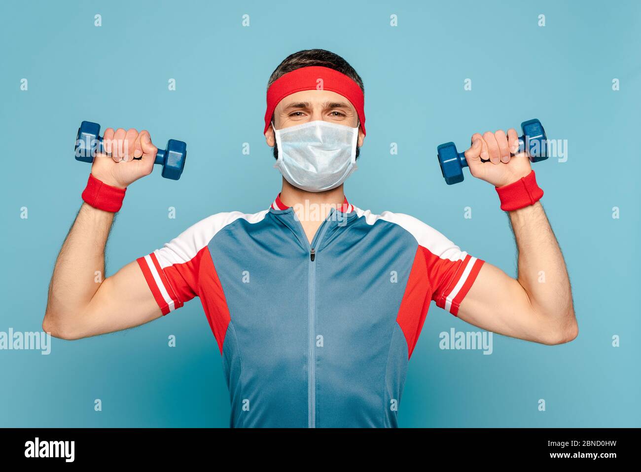 stylish sportsman in medical mask exercising with dumbbells on blue ...