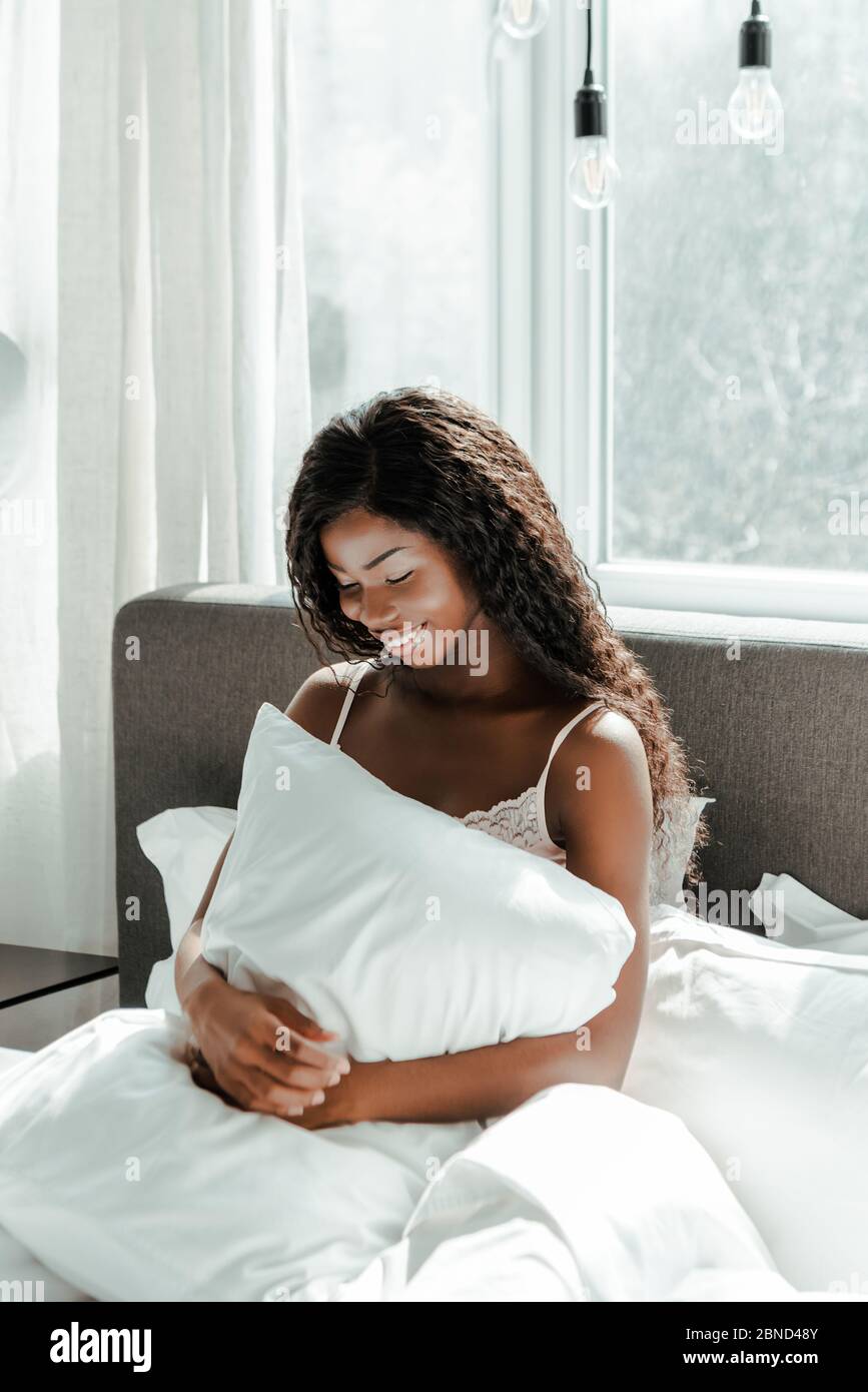 African american woman holding pillow and smiling on bed in bedroom ...