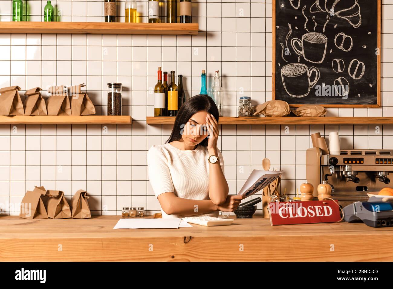 Stressed cafe owner with closed eyes holding papers near table with ...