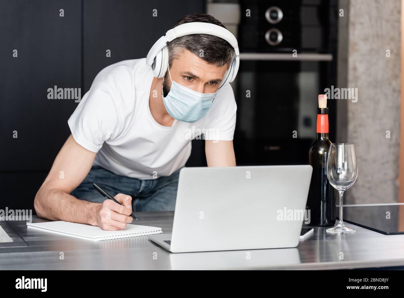 Teleworker in headphones and medical mask writing on notebook near ...