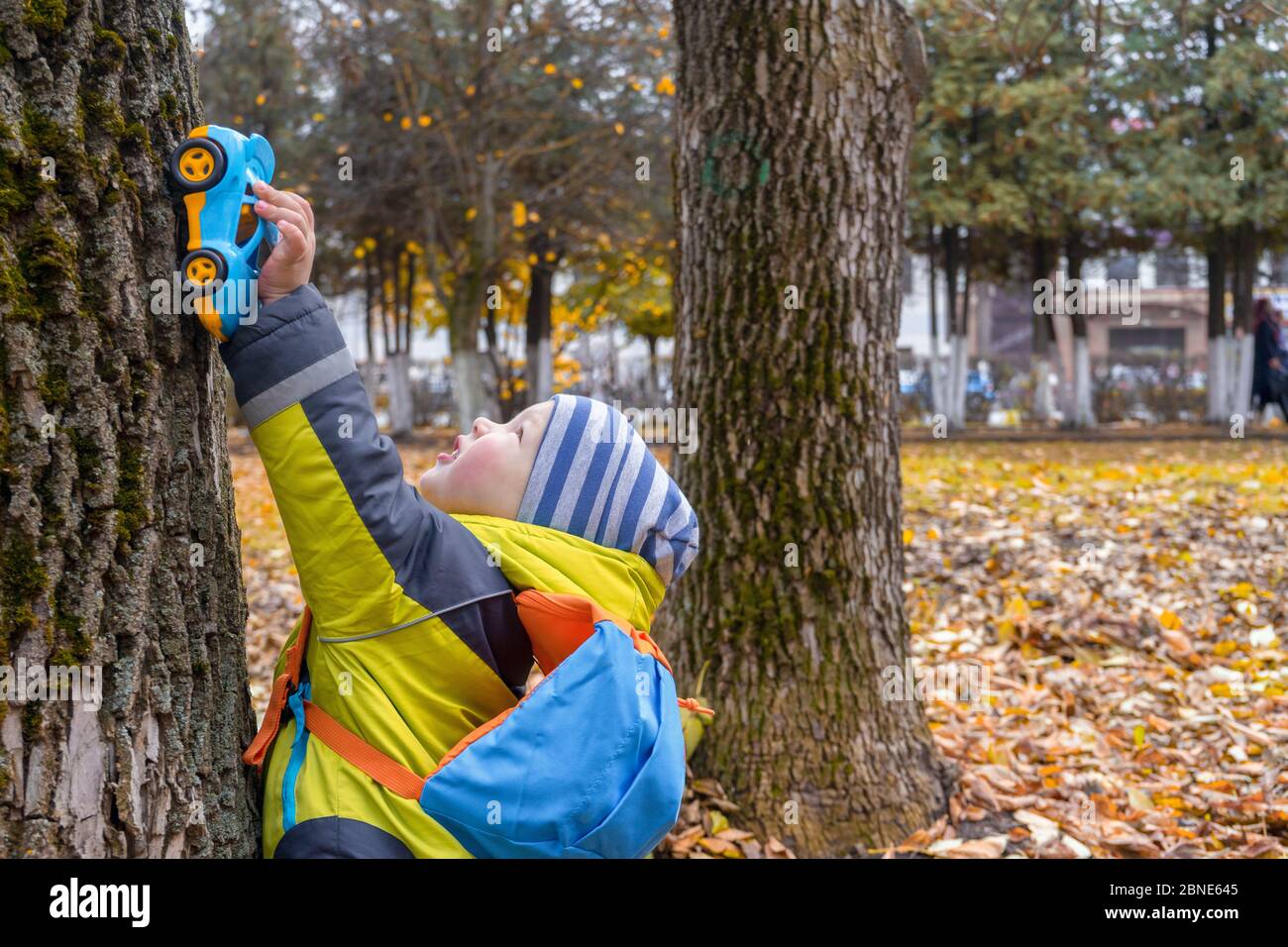 Little boy in yellow jacket, striped hat, with backpack plays with blue ...