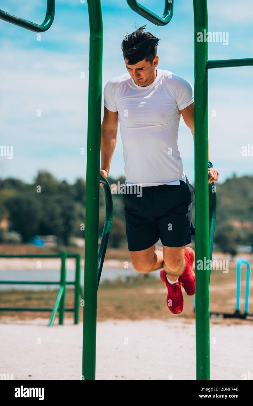 Strong muscular bodybuilder doing exercise on bars in the outdoors gym ...