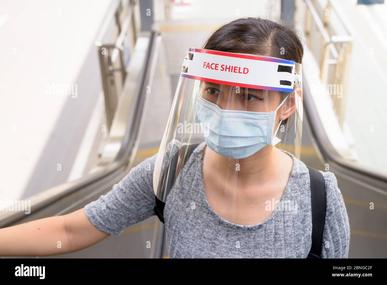 Young Asian woman with mask and face shield moving up the escalator ...