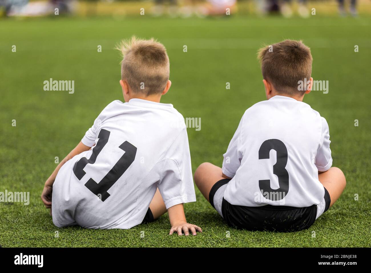 Two boys in sports uniforms sitting on grass pitch. Kids playing team ...