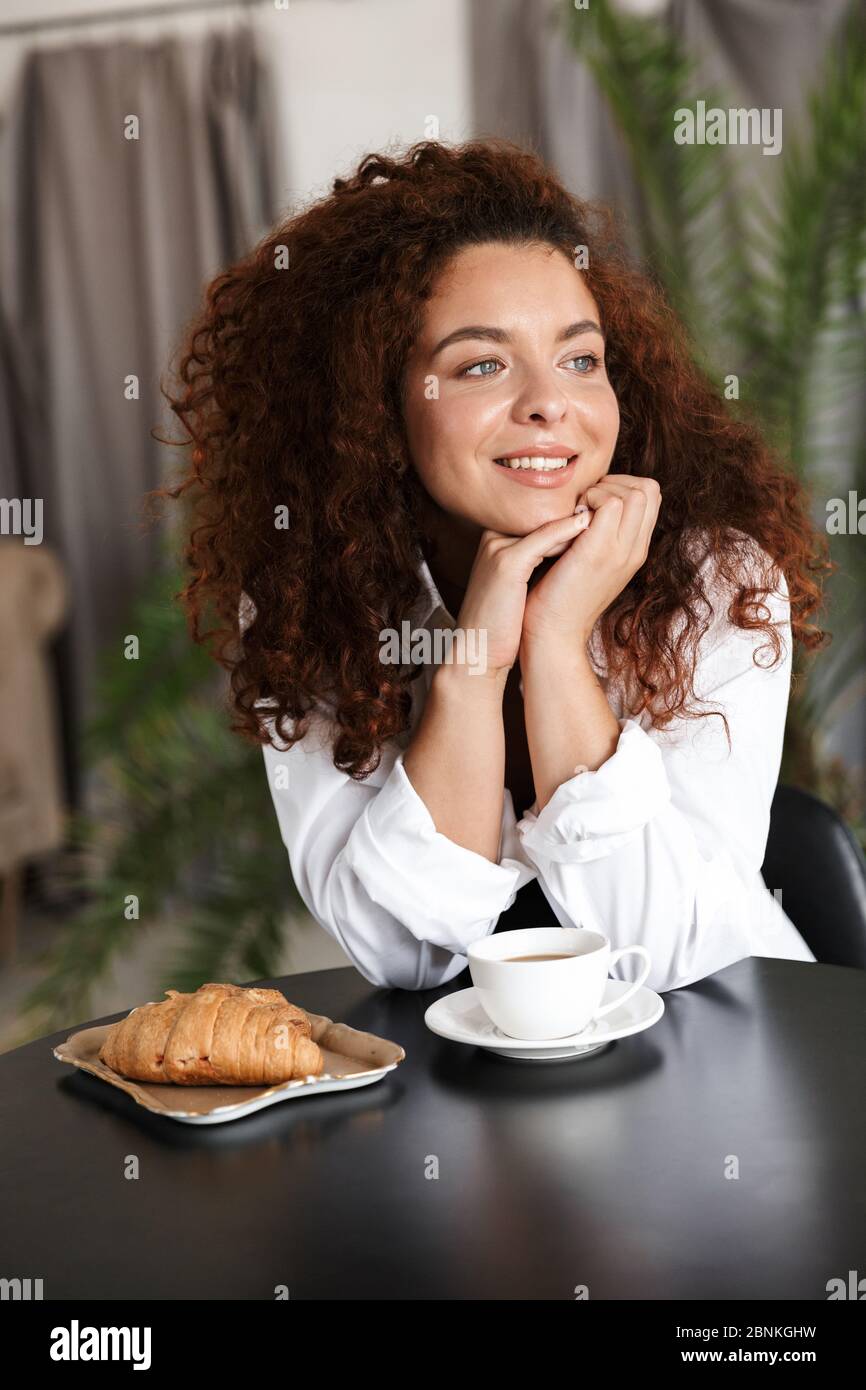Image of a smiling beautiful optimistic young woman dressed in white shirt indoors in home hotel ...