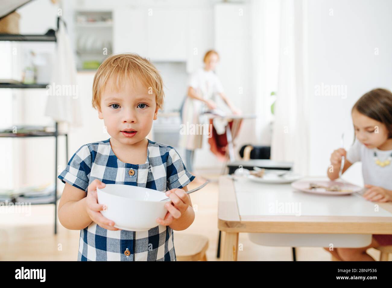 Portrait of toddler boy standing with bowl in hands, sister eating, mom ...