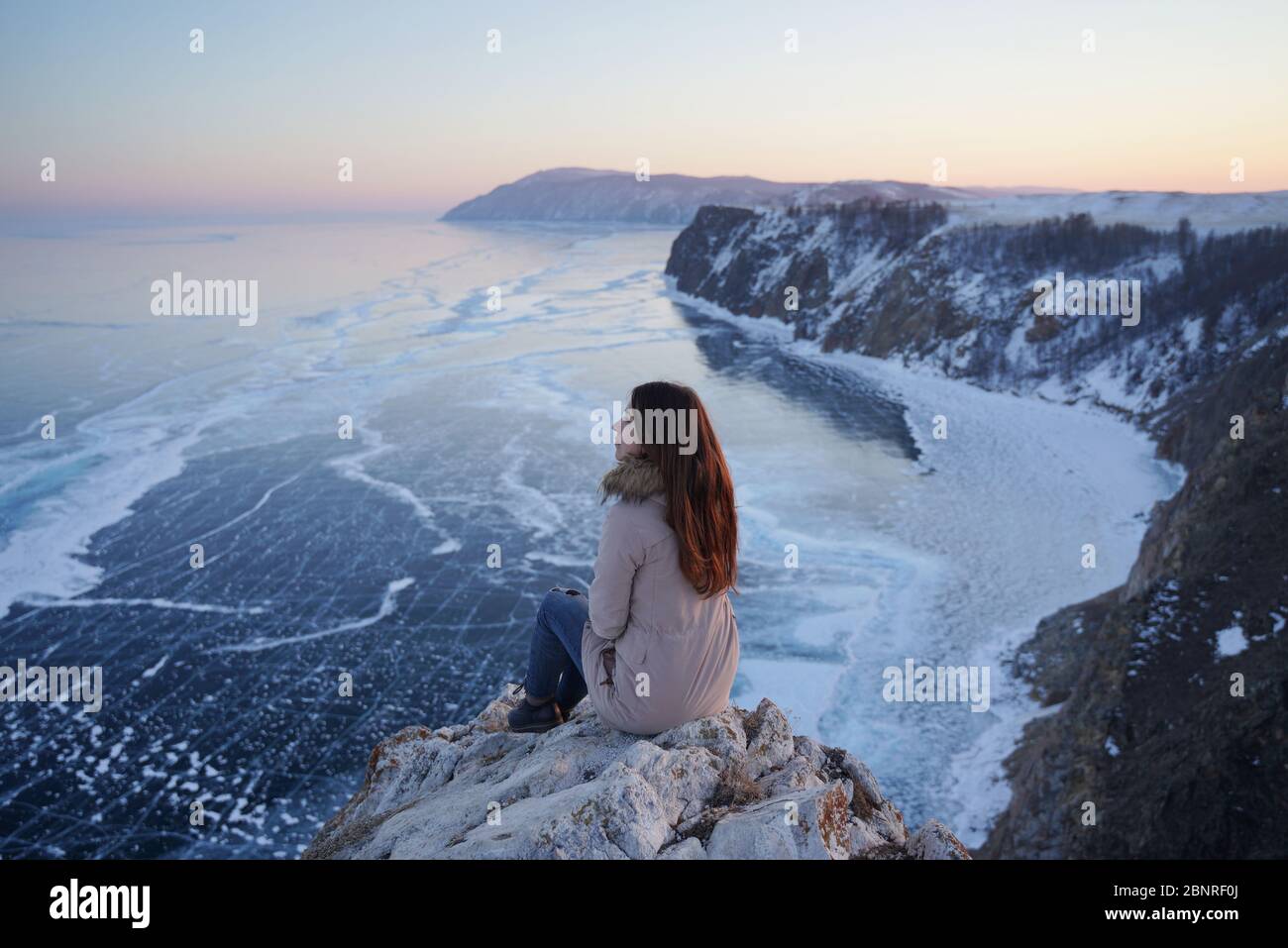 Lake Baikal at winter. Woman sitting on a cliff and looking at frozen Baikal lake. Deepest and ...