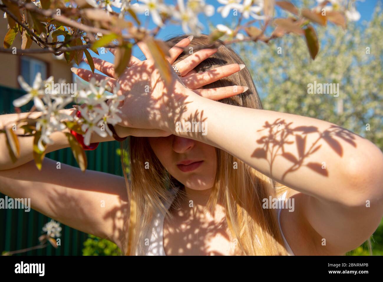 A teenage girl stands by a flowering shrub with her face in her hands ...