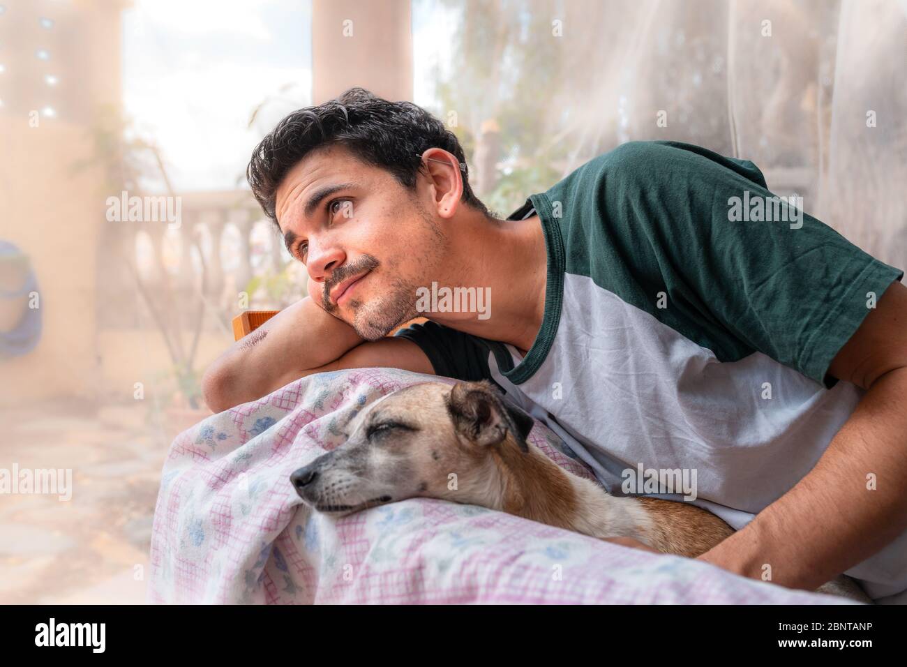 young man hugs a dog lying on an armchair in the garden, and smiles ...