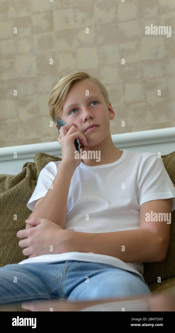Young blond handsome boy thinking while talking on the phone at home ...