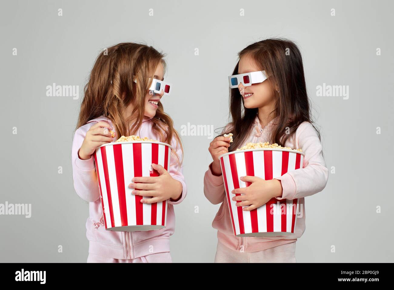 two beautiful caucasian little girls wearing red-blue 3d glasses and eating popcorn from bucket ...