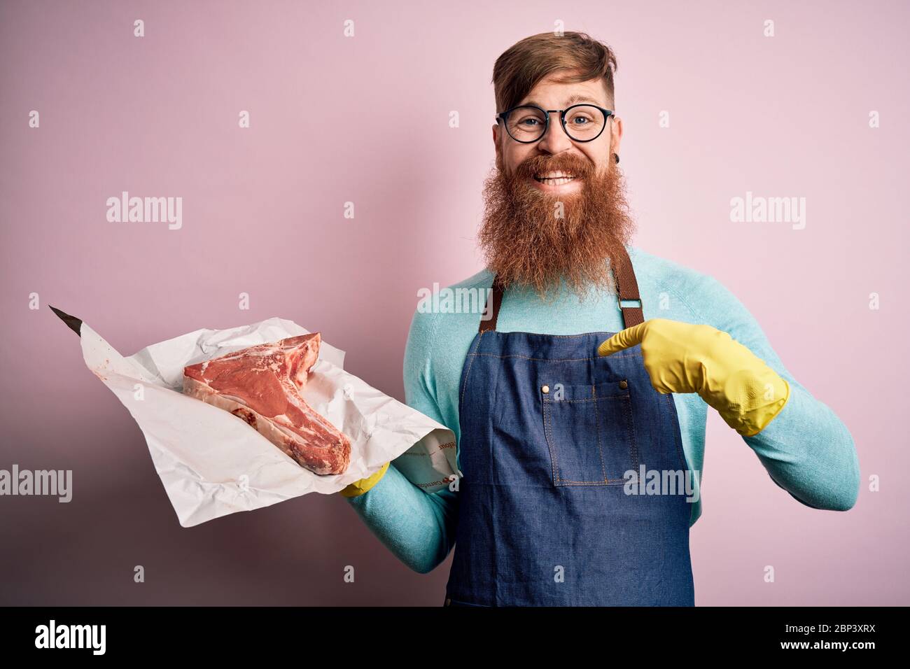 Redhead Irish butcher man with beard holding raw beef steak over pink ...
