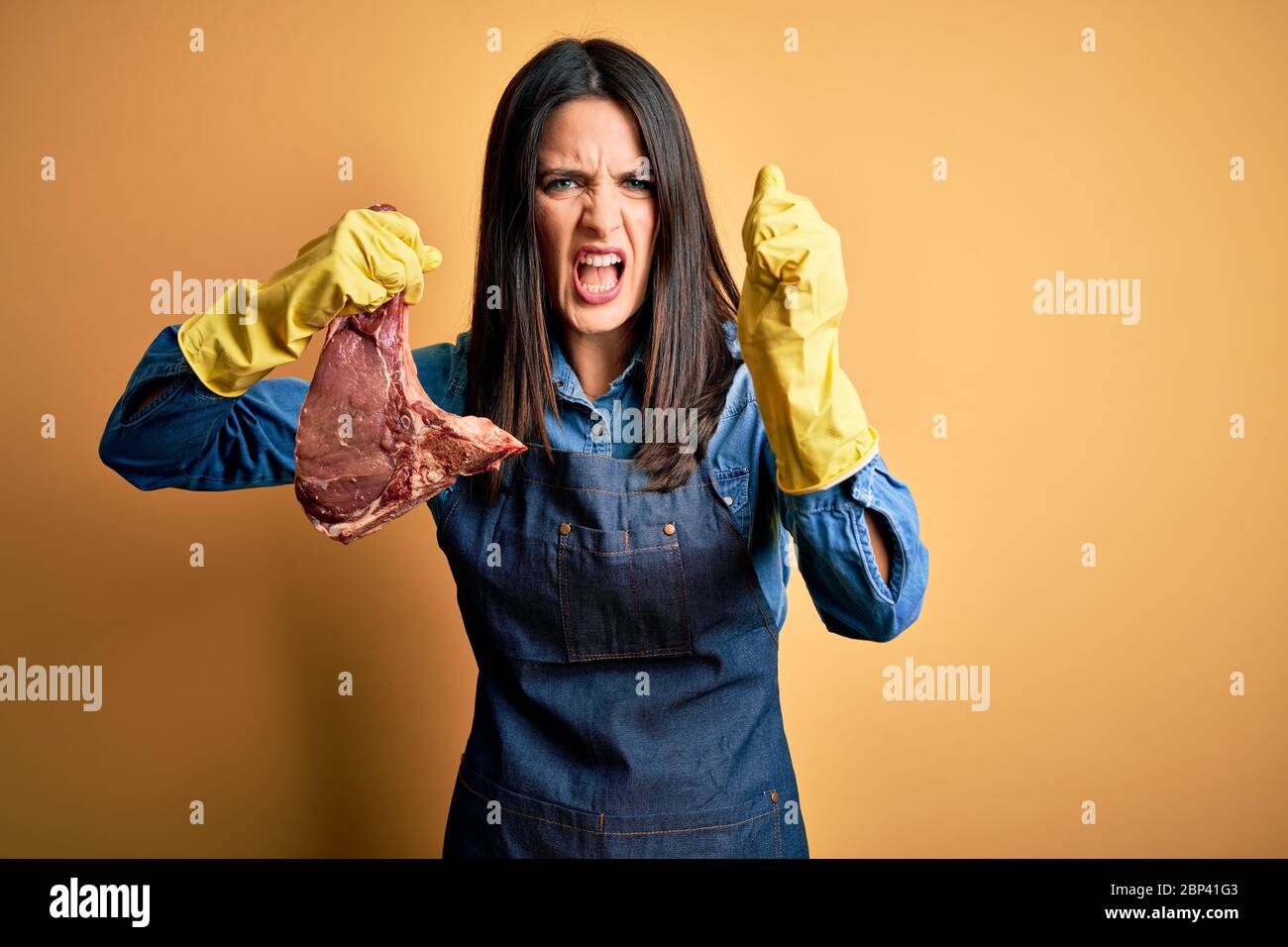 Young butcher woman wearing apron holding raw beef steak over yellow ...