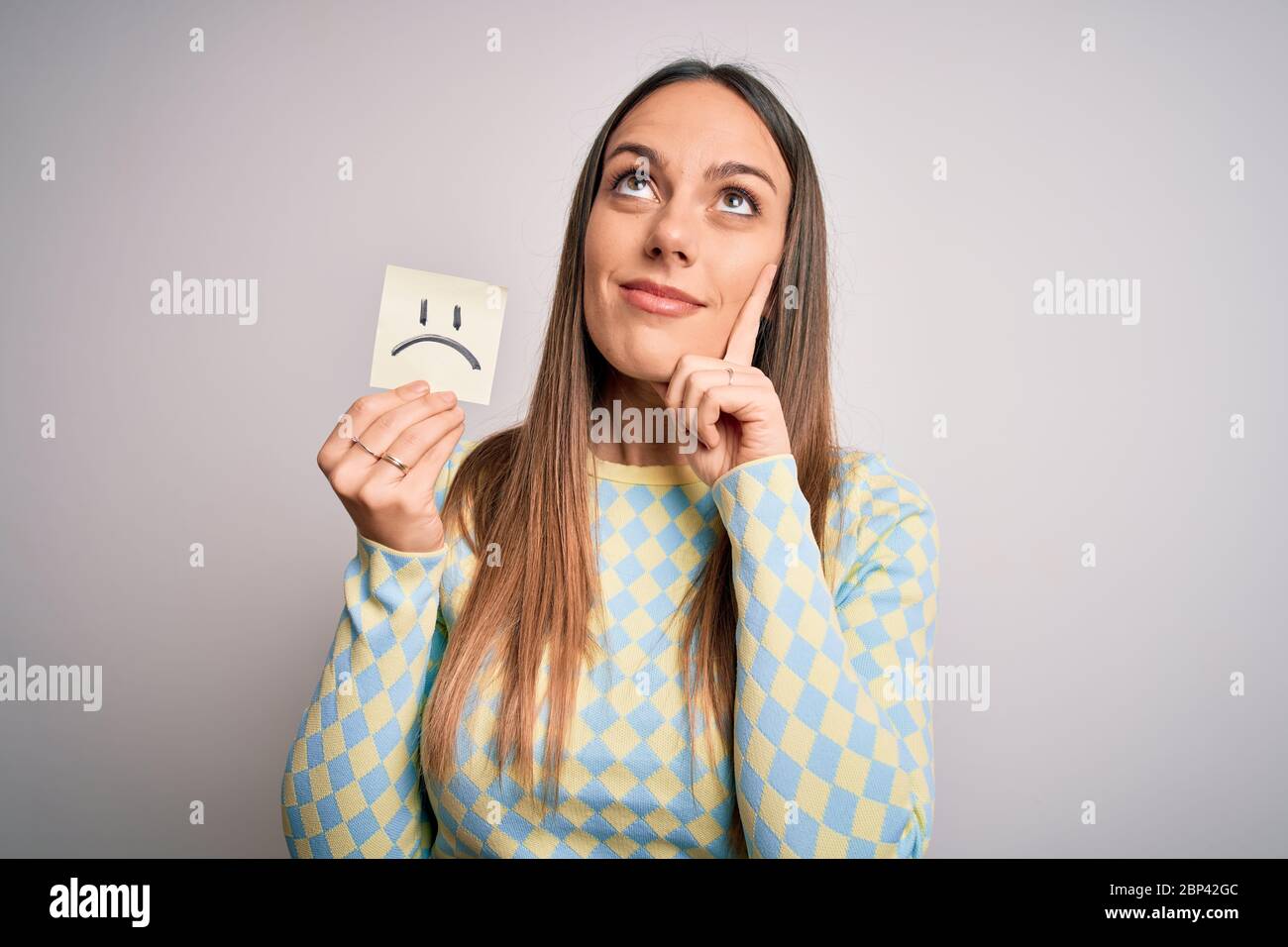 Young blonde woman holding sticky paper note with sad face emoticon ...