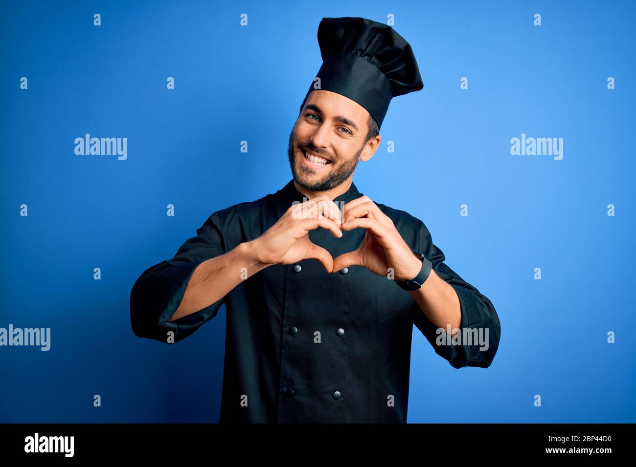 Young handsome chef man with beard wearing cooker uniform and hat over ...