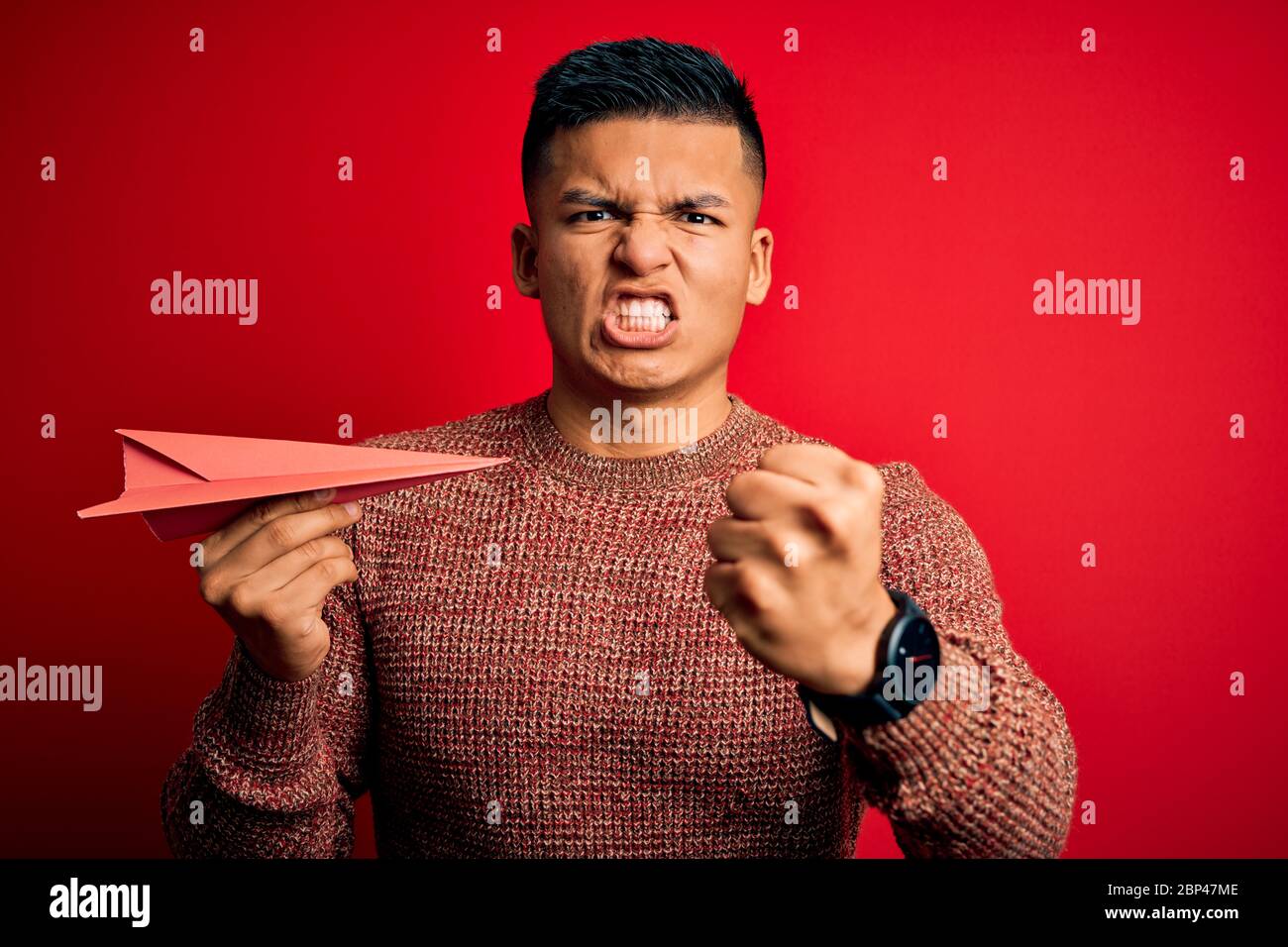 Young handsome latin man holding paper plane over isolated red ...