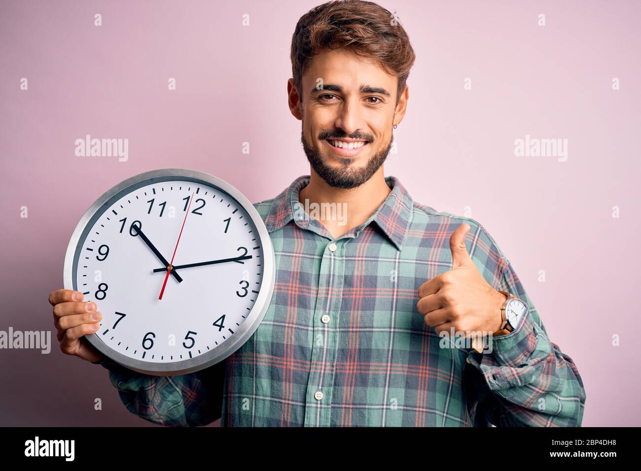 Young man with beard doing countdown using big clock over isolated pink ...