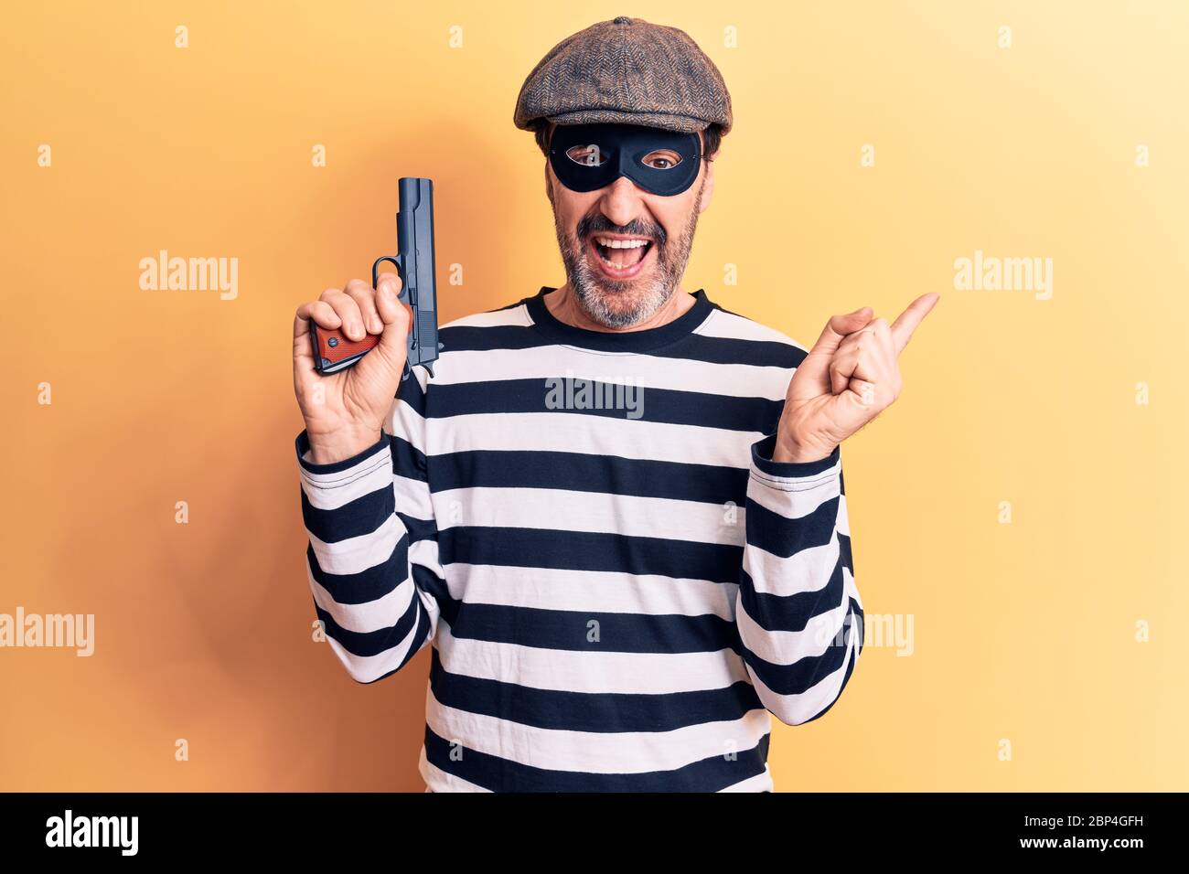 Middle age handsome burglar man wearing cap and mask holding gun over ...