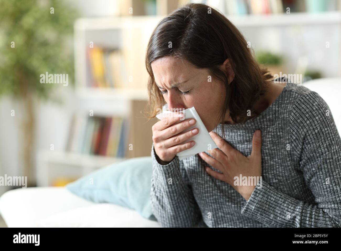 Sick adult woman coughing covering mouth with tissue sitting on a couch ...