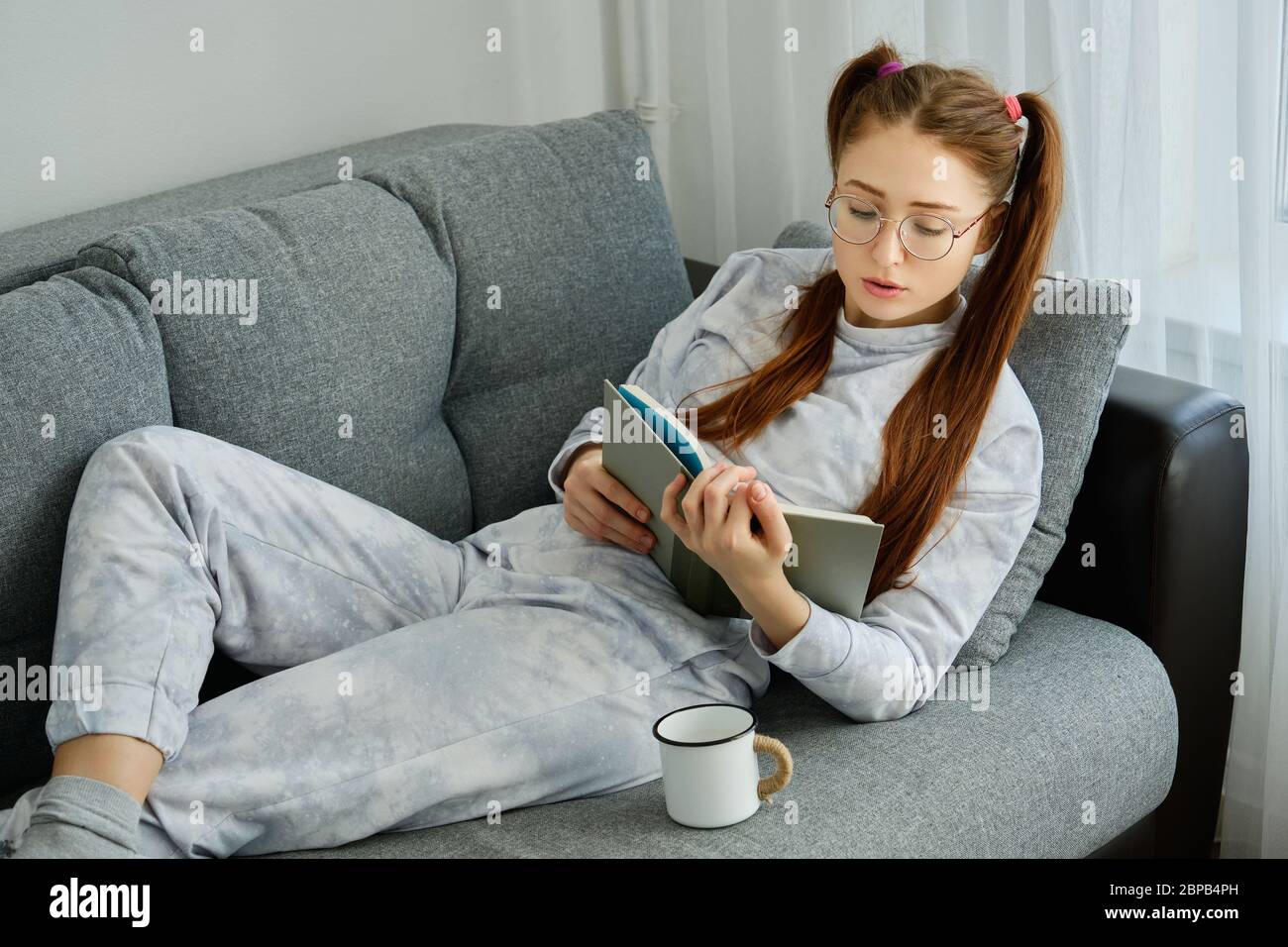 Red-haired girl with tails in pajamas lies on the sofa and reads a book ...