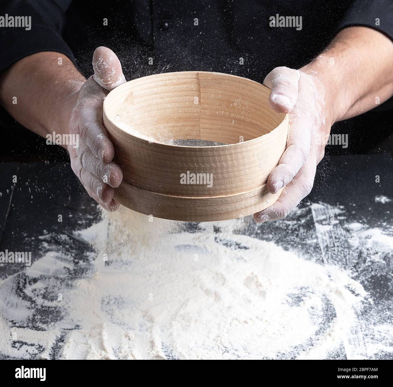 Chef in a black uniform holds in his hand a round wooden sieve and sift ...