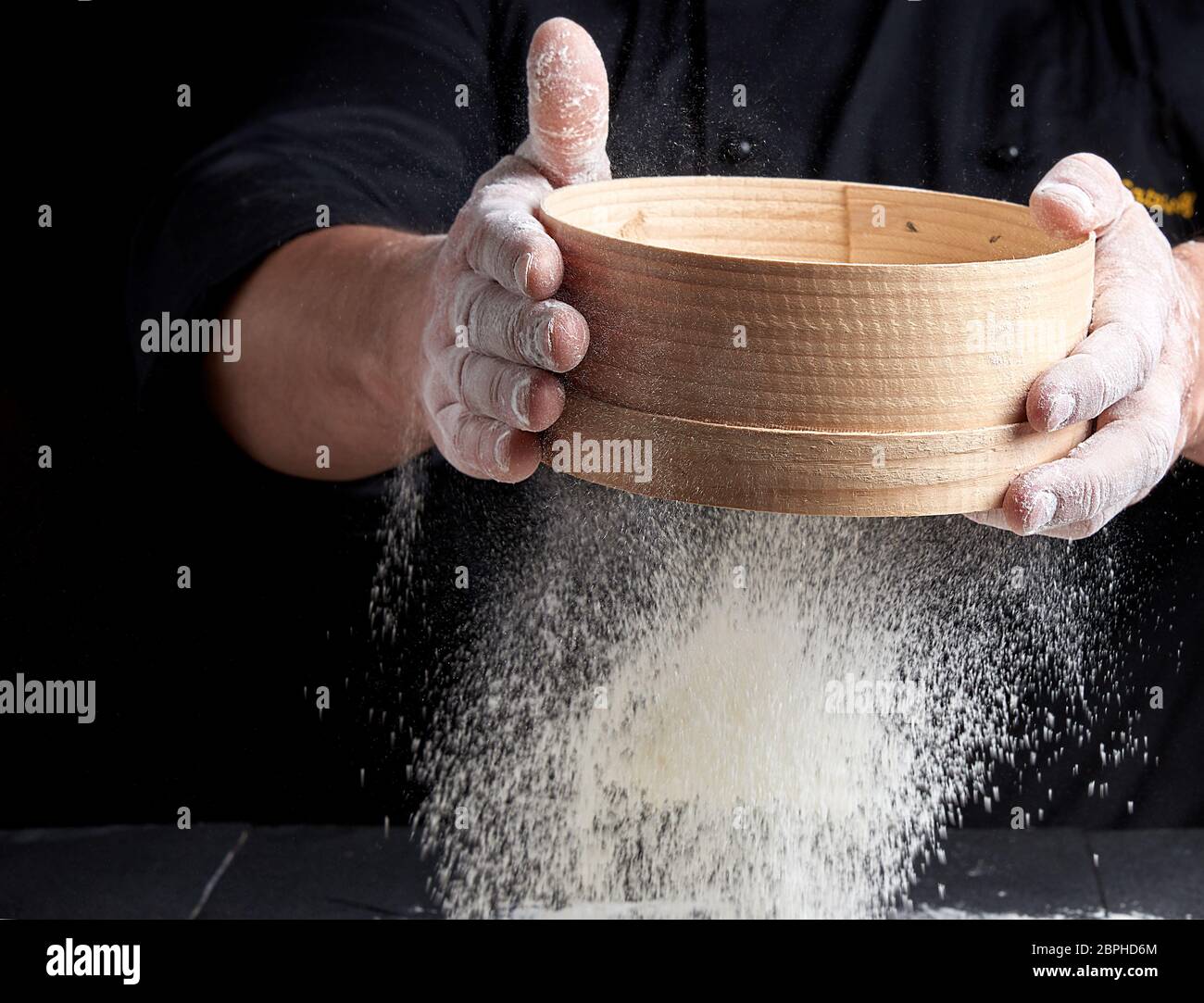 man sifts white wheat flour through a wooden sieve, black background ...