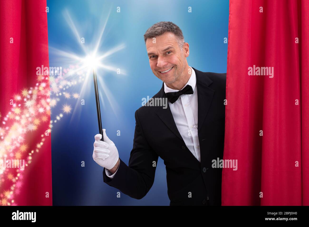 Happy Mature Male Magician Holding Magic Wand Behind Red Curtain Stock ...