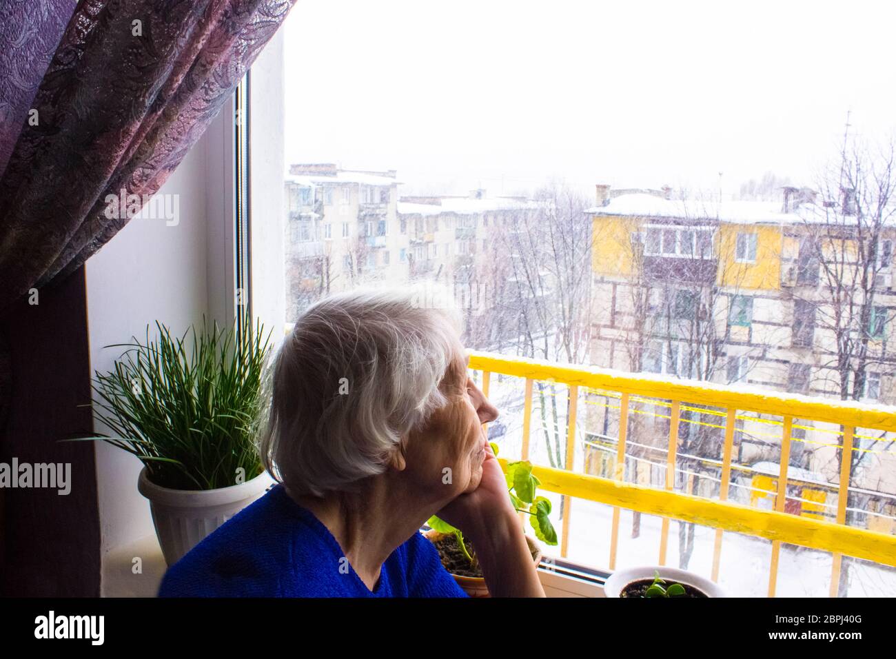 Old lonely woman sitting near the window in his house and looking at ...