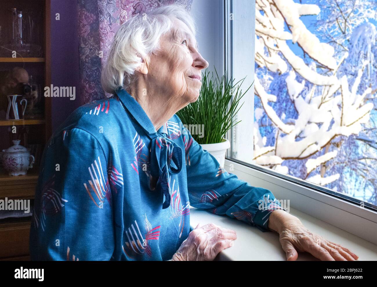 Old lonely woman sitting near the window in his house and looking and ...