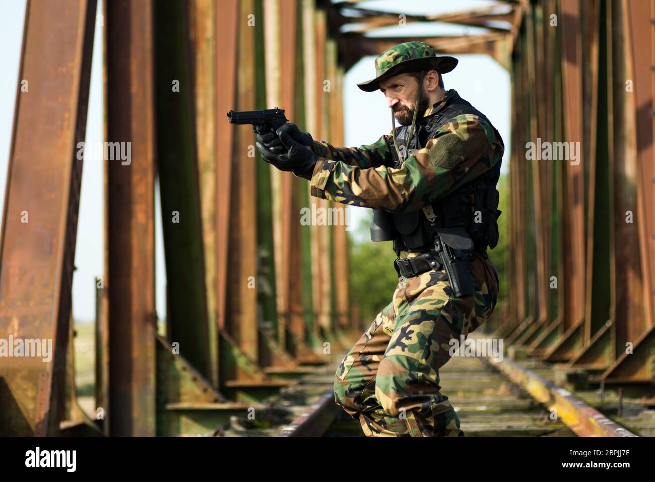 The soldier in military uniform is aiming with pistol on old railway ...
