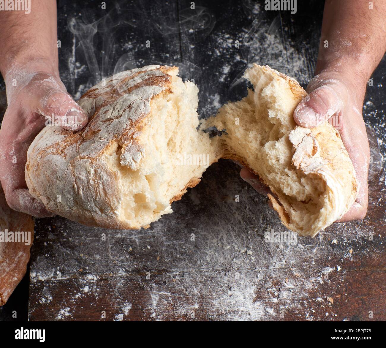 male hands breaking open baked bread in half over black wooden table ...