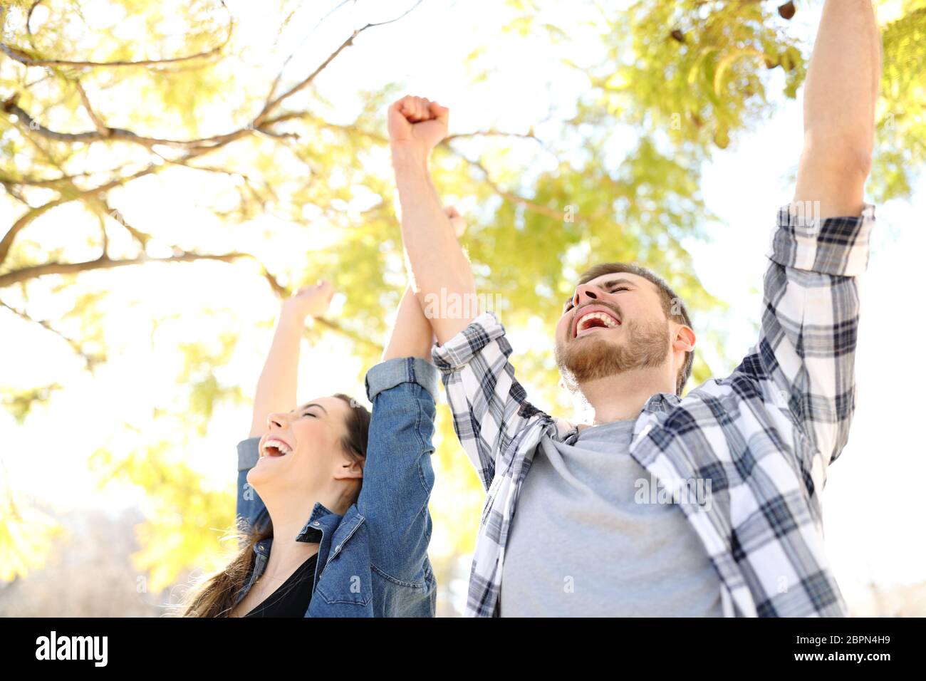 Excited couple celebrating success raising arms in a park with trees in ...