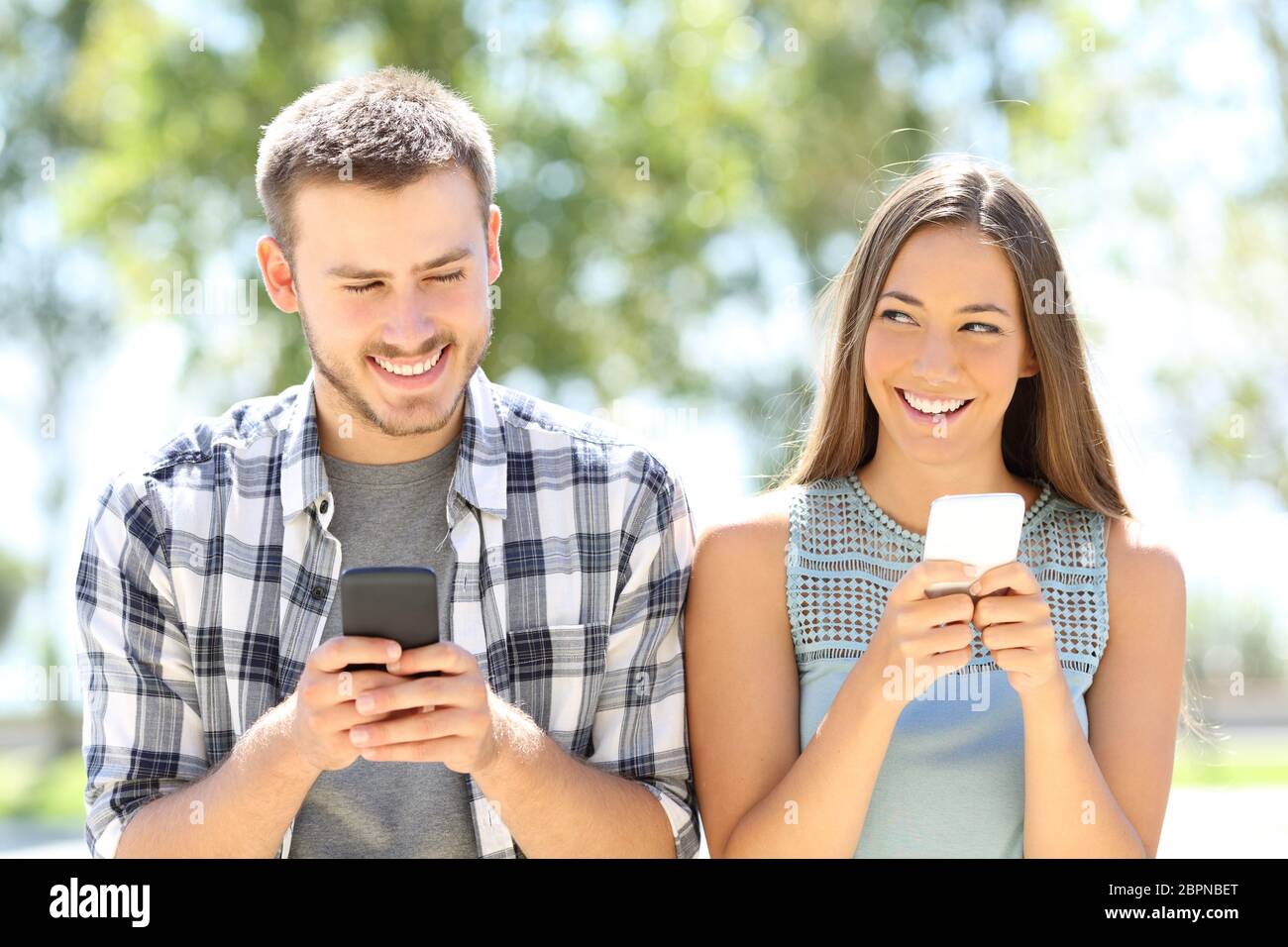 Front view of two friends flirting everyone with a smart phone and ...